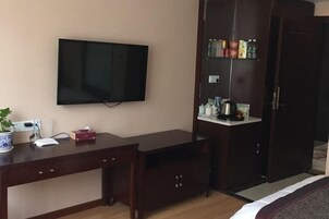 Quarto de hotel com cama, escrivaninha, televisão, guarda-roupa e prateleira com artigos de higiene pessoal.