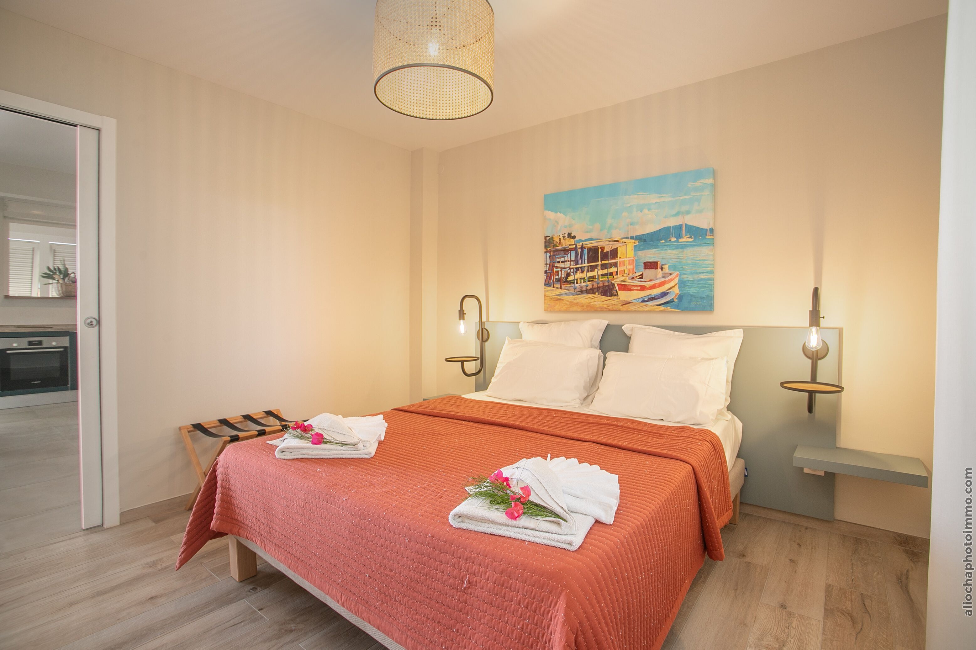 Cama bem arrumada com um cobertor vermelho, um quadro de um barco na parede e uma mesa pequena com uma luminária.