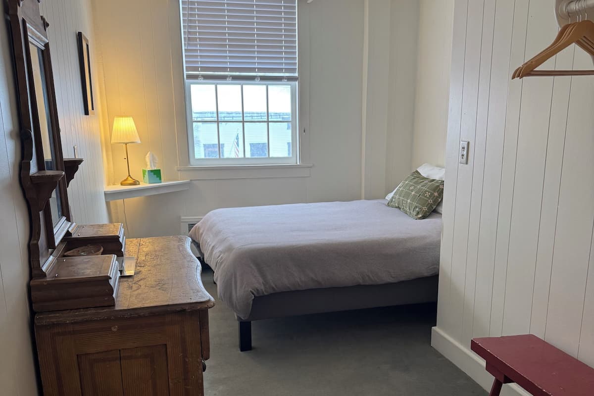 Quarto com cama, cômoda, janela, luminária e um cabideiro.