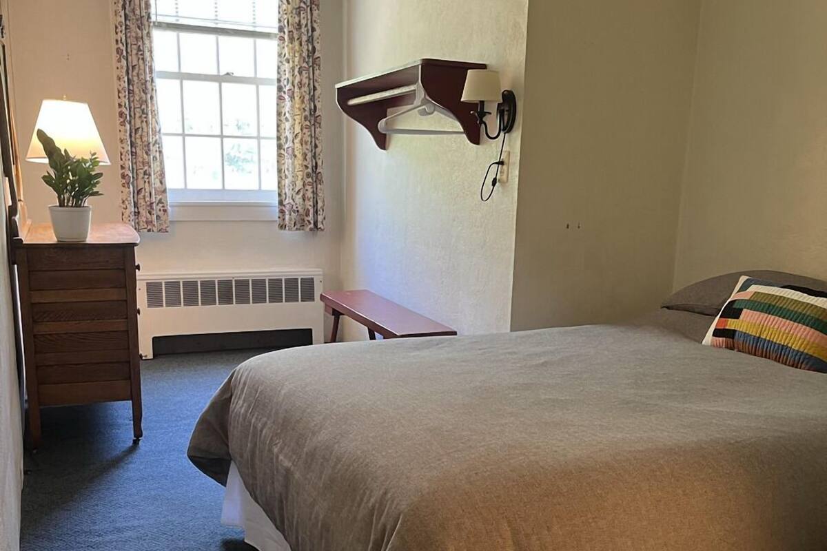 Um quarto com uma cama grande, uma mesa de cabeceira com abajur, um banco, um aquecedor na parede e uma janela com cortinas.