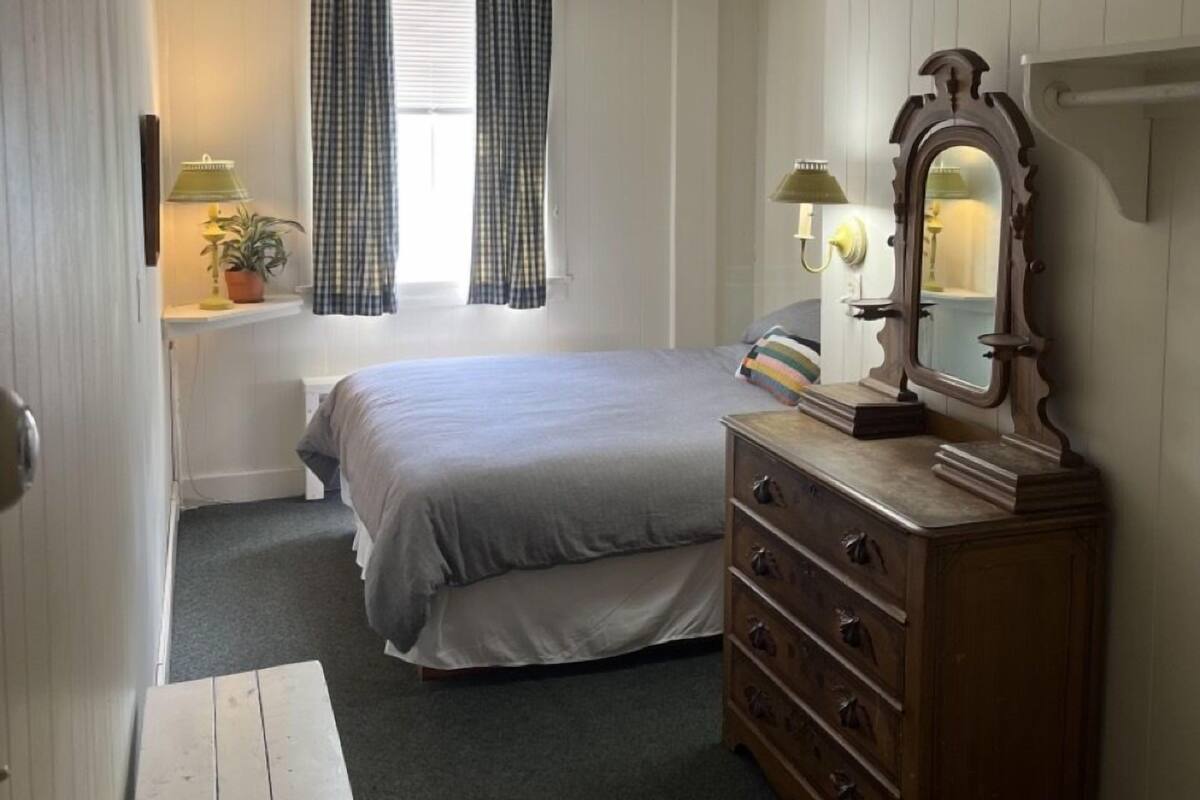 Quarto com cama, cómoda com espelho, janela com cortinas e candeeiro numa mesa de cabeceira.