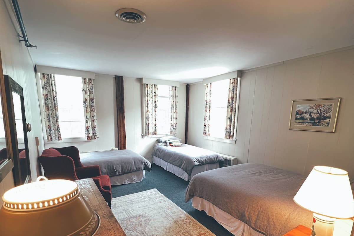 Quarto de hotel com duas camas de solteiro, uma escrivaninha de madeira com luminária, uma mesinha com luminária, um tapete e um quadro na parede.