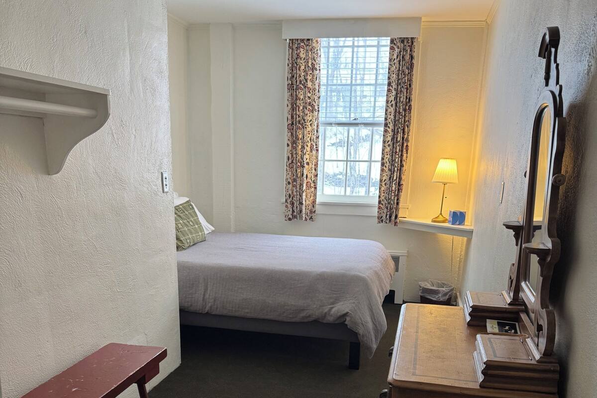 Um quarto com cama, um banco, cômoda, janela com cortinas e luminária.