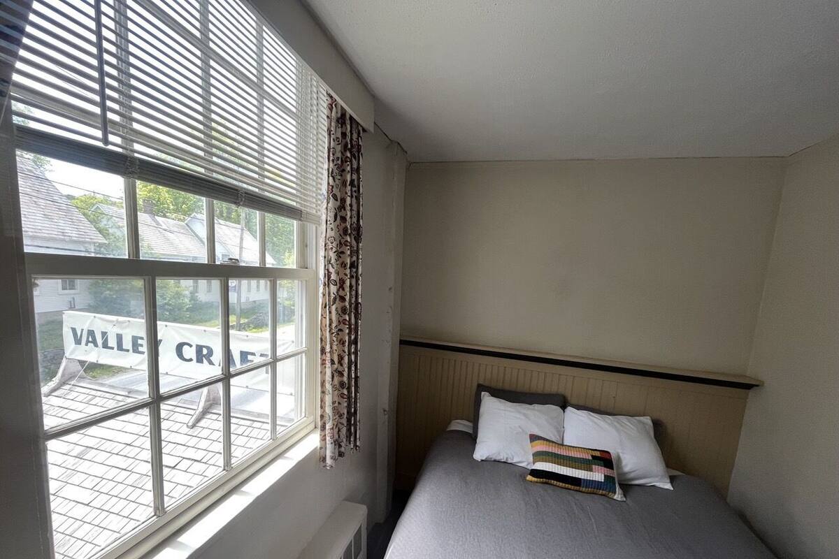 Quarto de hotel pequeno com uma cama de casal, janela com persianas e vista para uma placa de rua.