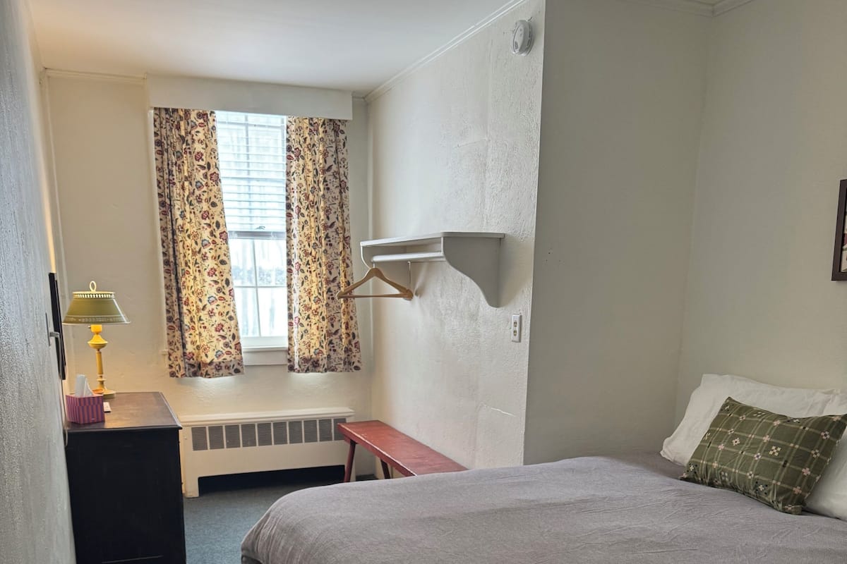 Quarto com cama, mesa de cabeceira com abajur, um banco, janela com cortinas e prateleira fixada na parede.