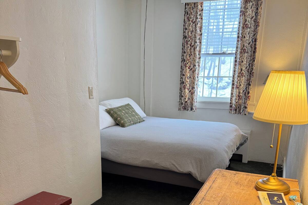 Quarto pequeno com uma cama de solteiro, mesa de cabeceira de madeira, um banco, janela com cortinas florais e aquecedor fixado na parede.