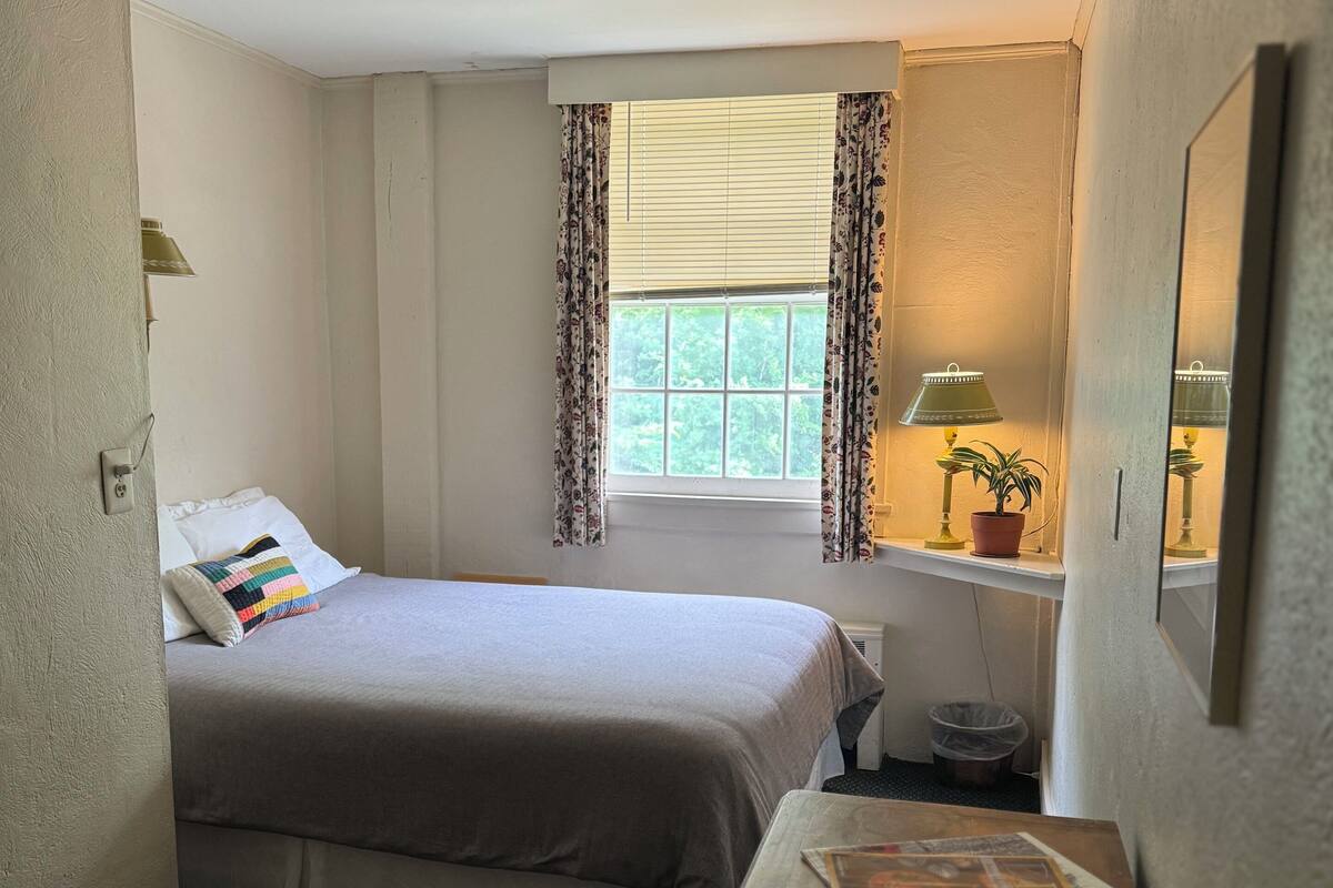 Quarto de hotel com cama, mesa de cabeceira, abajur, janela com cortinas e luminária na parede.