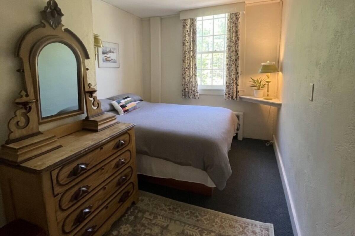 Um quarto com cama, cómoda, espelho, janela com cortinas e uma luminária.
