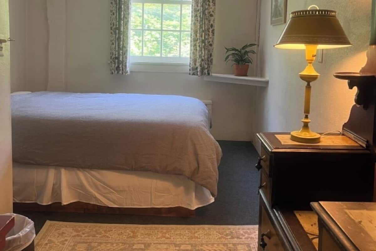 Quarto com cama, janela com cortinas, luminária, cômoda e tapete.