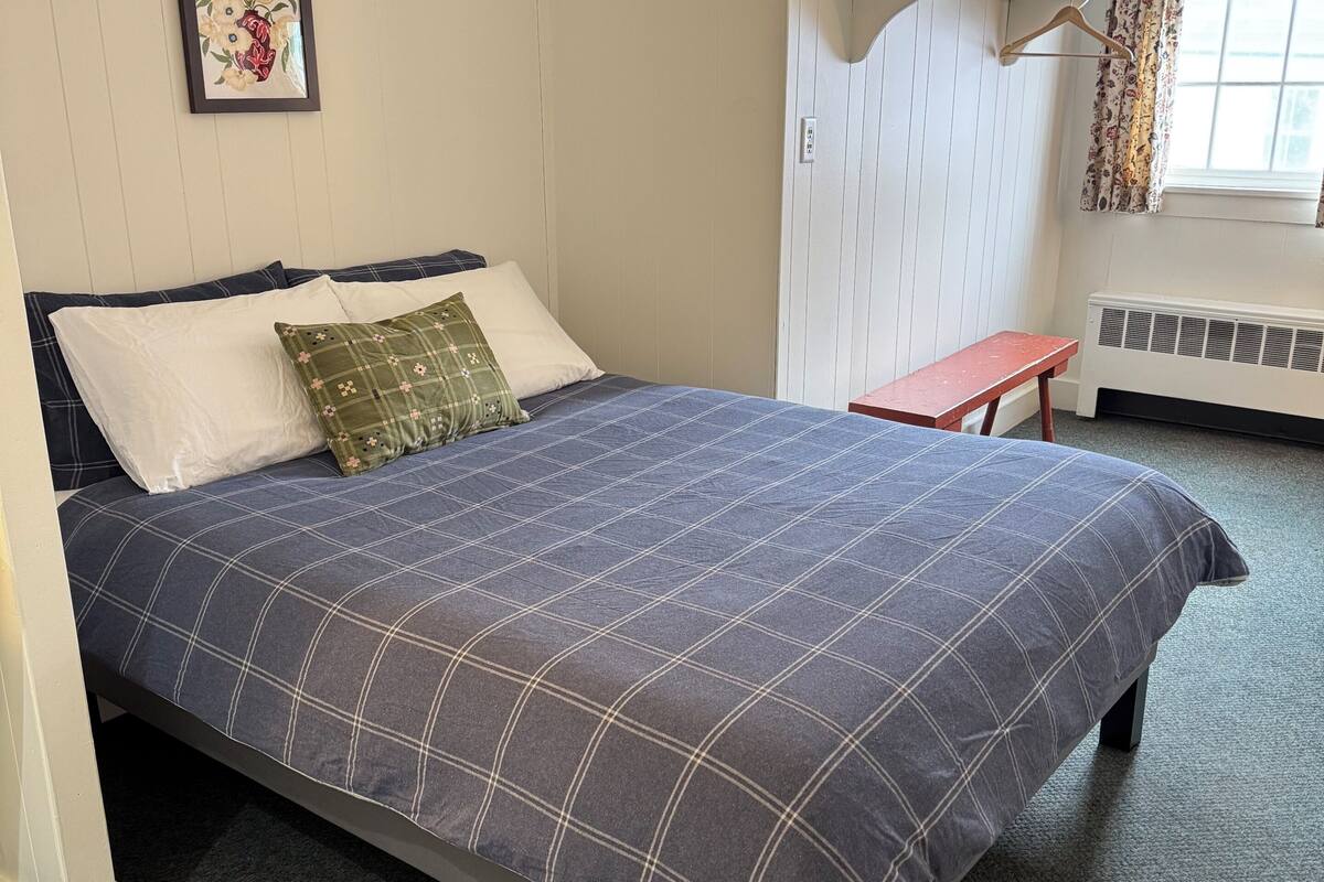 Quarto com cama, almofadas, um quadro emoldurado, uma mesa pequena e uma janela com cortinas.