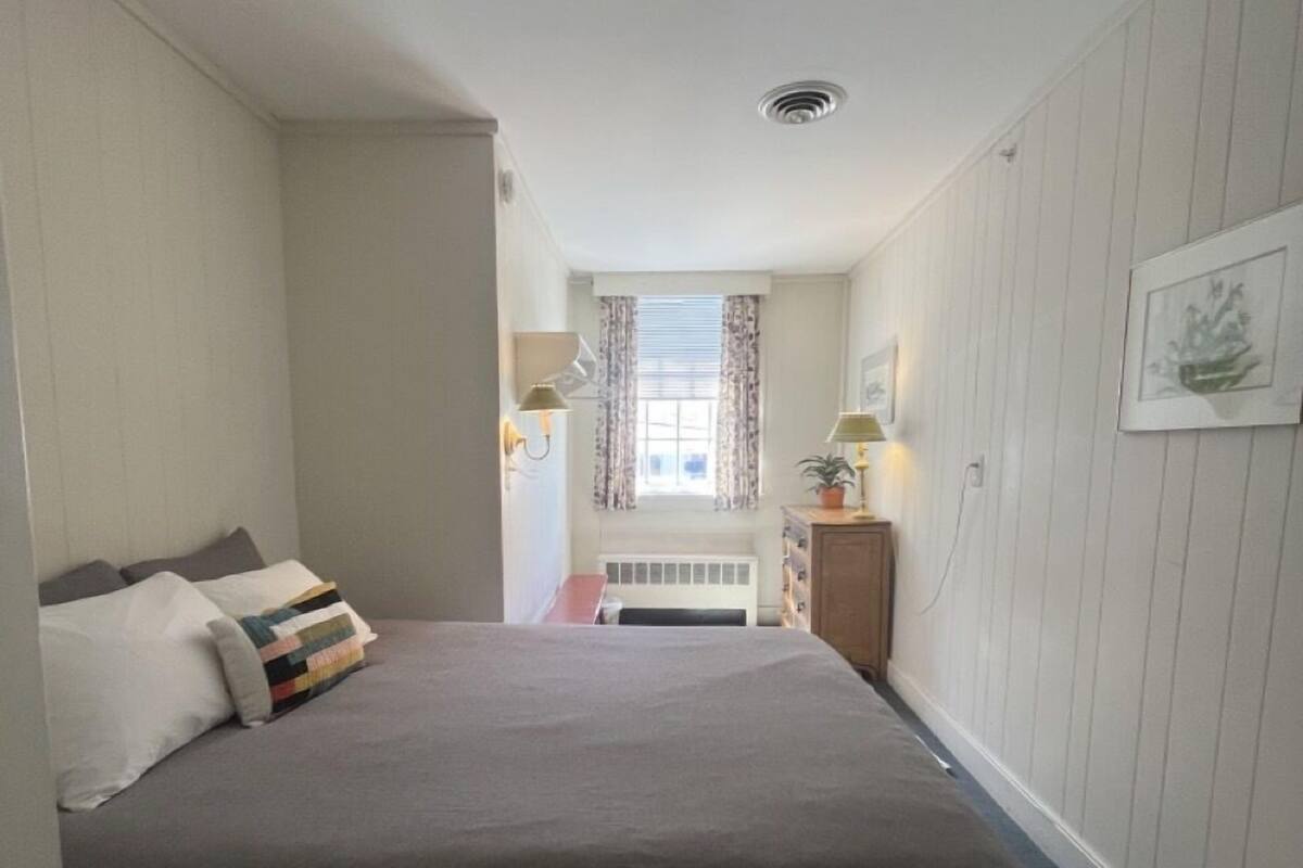 Um quarto com uma cama grande, uma mesa de cabeceira com abajur, uma janela com cortinas e um radiador.