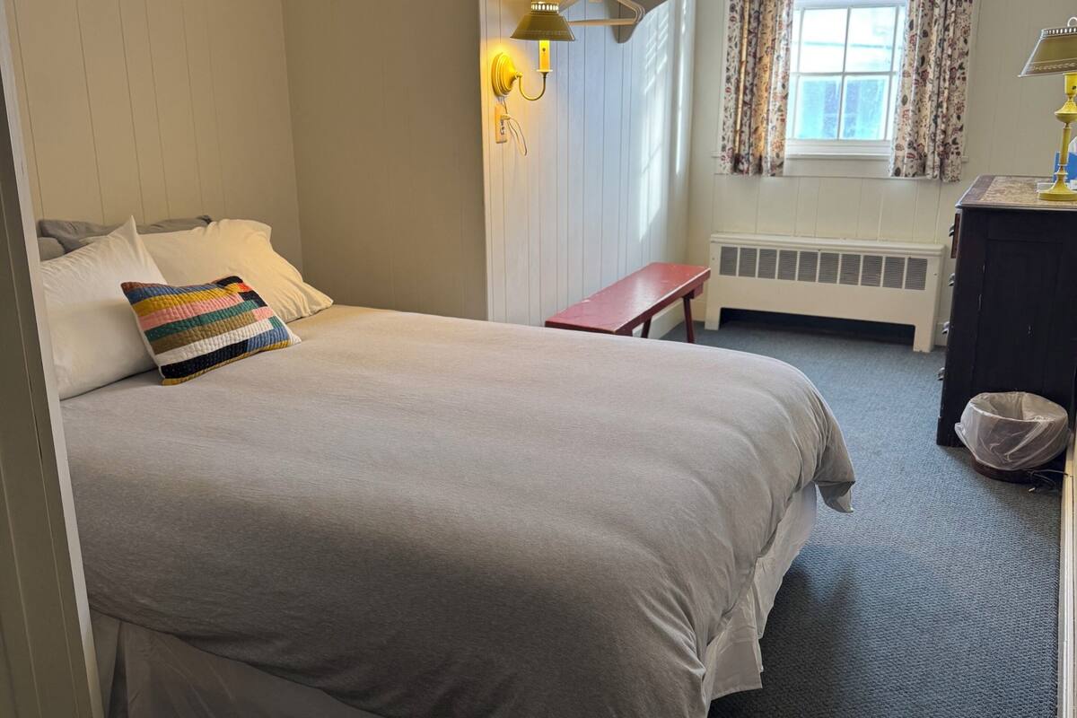 Quarto de hotel com cama, um banco, escrivaninha, abajur e janela com cortinas.