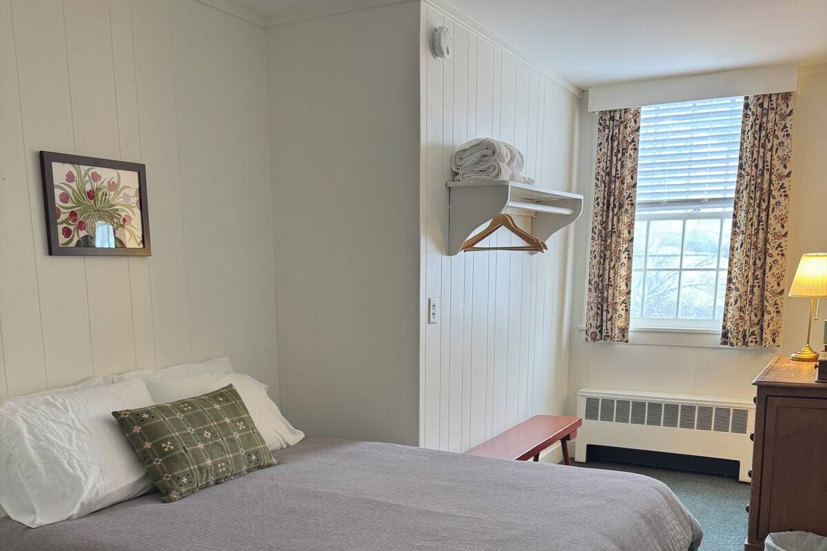 Quarto com cama, mesa de cabeceira, luminária, janela com cortinas e aquecedor fixado na parede.