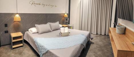 Quarto de hotel moderno com uma cama grande, mesas de cabeceira, um banco de madeira e uma televisão fixada na parede.