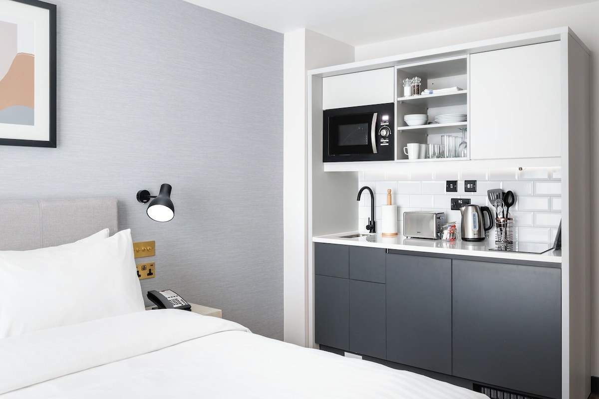 Quarto de hotel com cama, mesa de cabeceira com abajur, telefone, micro-ondas, máquina de café e pequena cozinha com armários e pia.