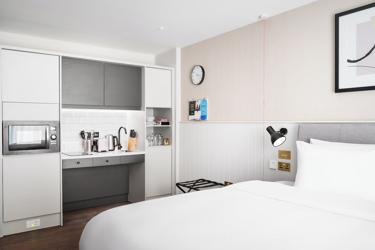 Quarto de hotel moderno com cozinha compacta, uma cama, um relógio de parede e um quadro emoldurado.