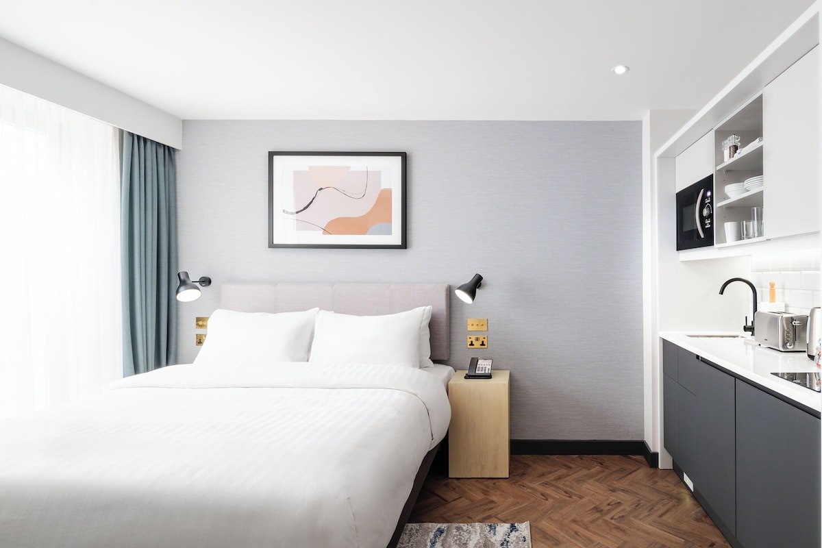 Quarto de hotel moderno com cama, mesa de cabeceira, luminária, obra de arte e uma cozinha compacta com pia e micro-ondas.