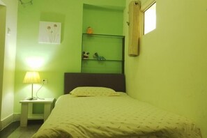 Um quarto pequeno e bem arrumado, com uma cama, uma mesa de cabeceira com abajur, uma estante com brinquedos e uma janela.