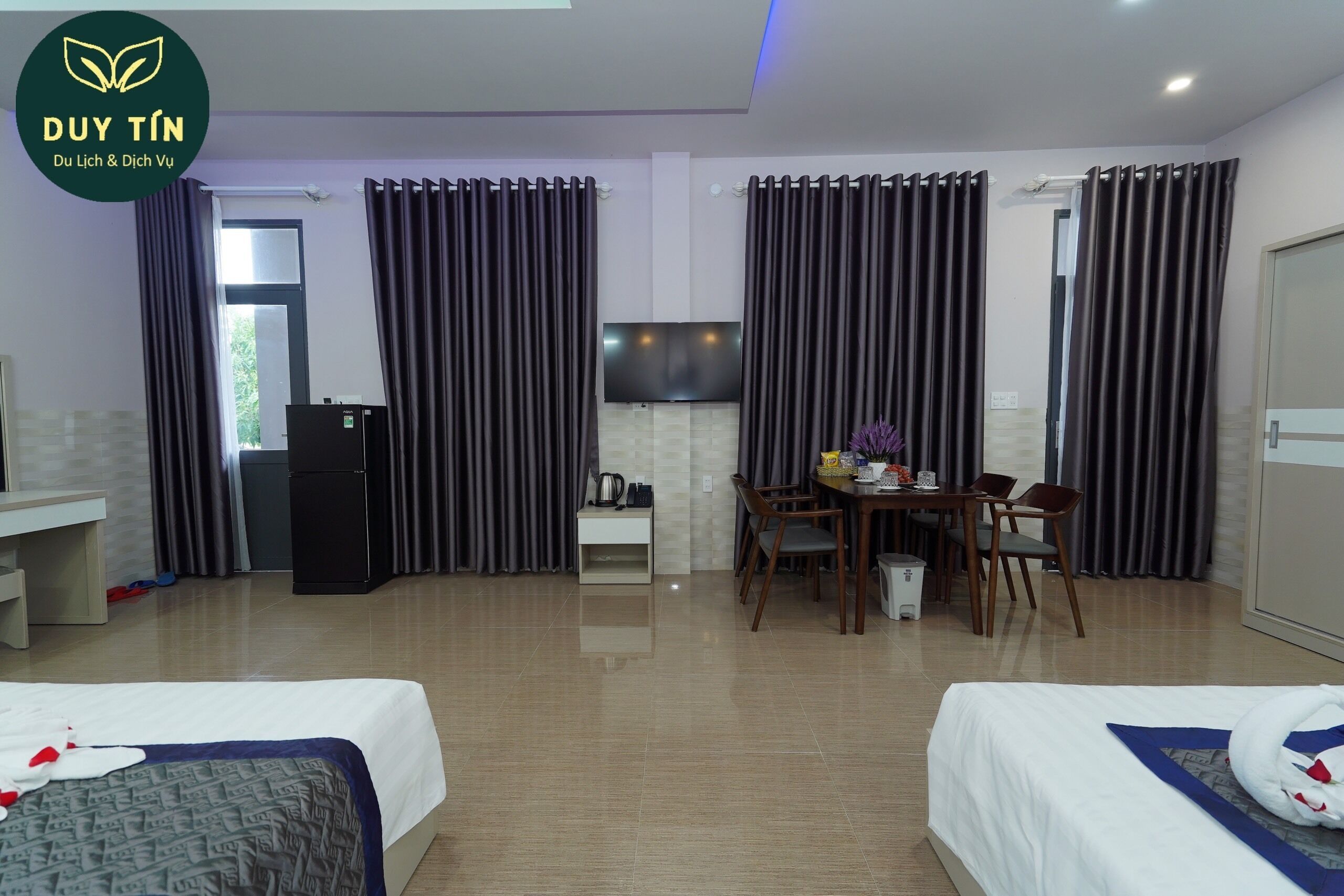 Quarto de hotel com duas camas, uma mesa de jantar com cadeiras, televisão e uma área de cozinha compacta.