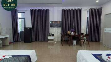 Quarto de hotel com duas camas, uma mesa de jantar com cadeiras, televisão e uma área de cozinha compacta.