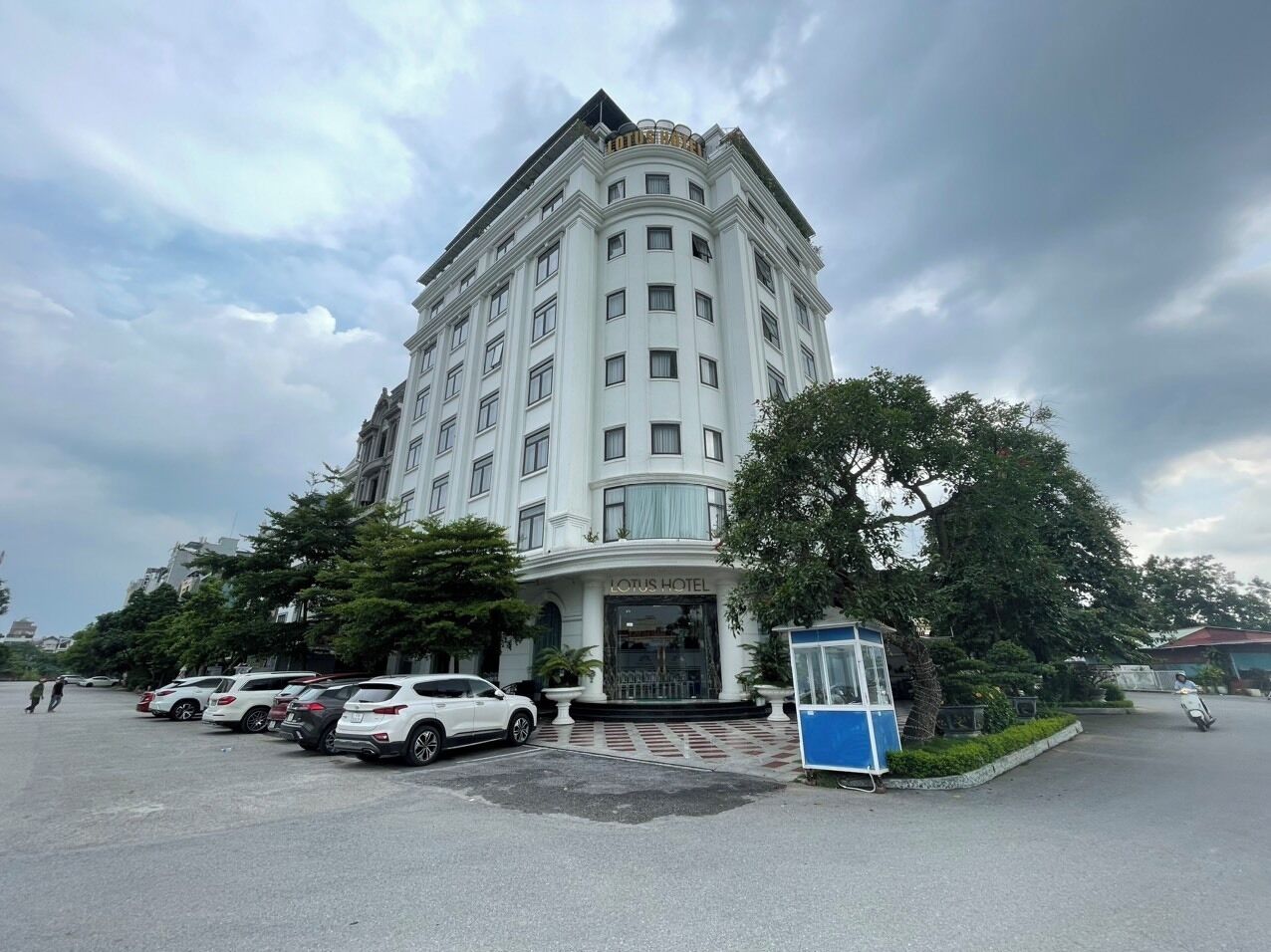 Um hotel de vários andares com uma entrada imponente, um estacionamento com vários carros e uma pequena estrutura azul perto da entrada.