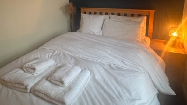 Uma cama bem arrumada com lençóis e toalhas brancas, uma cabeceira de madeira e um abajur.