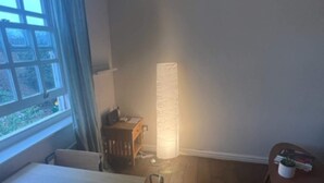 Quarto com piso de madeira, uma mesa, uma cadeira, uma luminária e uma janela com cortinas.