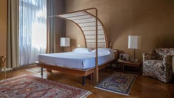 Quarto com uma cama de dossel curvada, duas luminárias de apoio, uma poltrona estampada e um tapete com padrão.