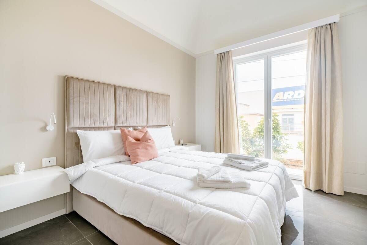 Quarto de hotel com uma cama grande, cabeceira, mesa de cabeceira e vista para o exterior pela janela.