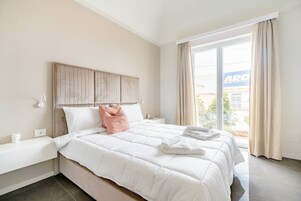 Quarto de hotel com uma cama grande, cabeceira, mesa de cabeceira e vista para o exterior pela janela.