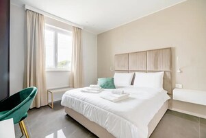 Quarto de hotel moderno com uma cama grande, uma mesa de cabeceira, uma cadeira e uma janela com cortinas.