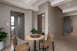 Quarto de hotel moderno com uma mesa de jantar redonda, duas cadeiras de vime, uma planta em vaso e vista da cidade pela porta.