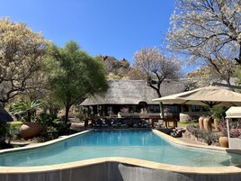 Piscina com espreguiçadeiras e guarda-sóis, cercada por árvores e um edifício com telhado de palha.