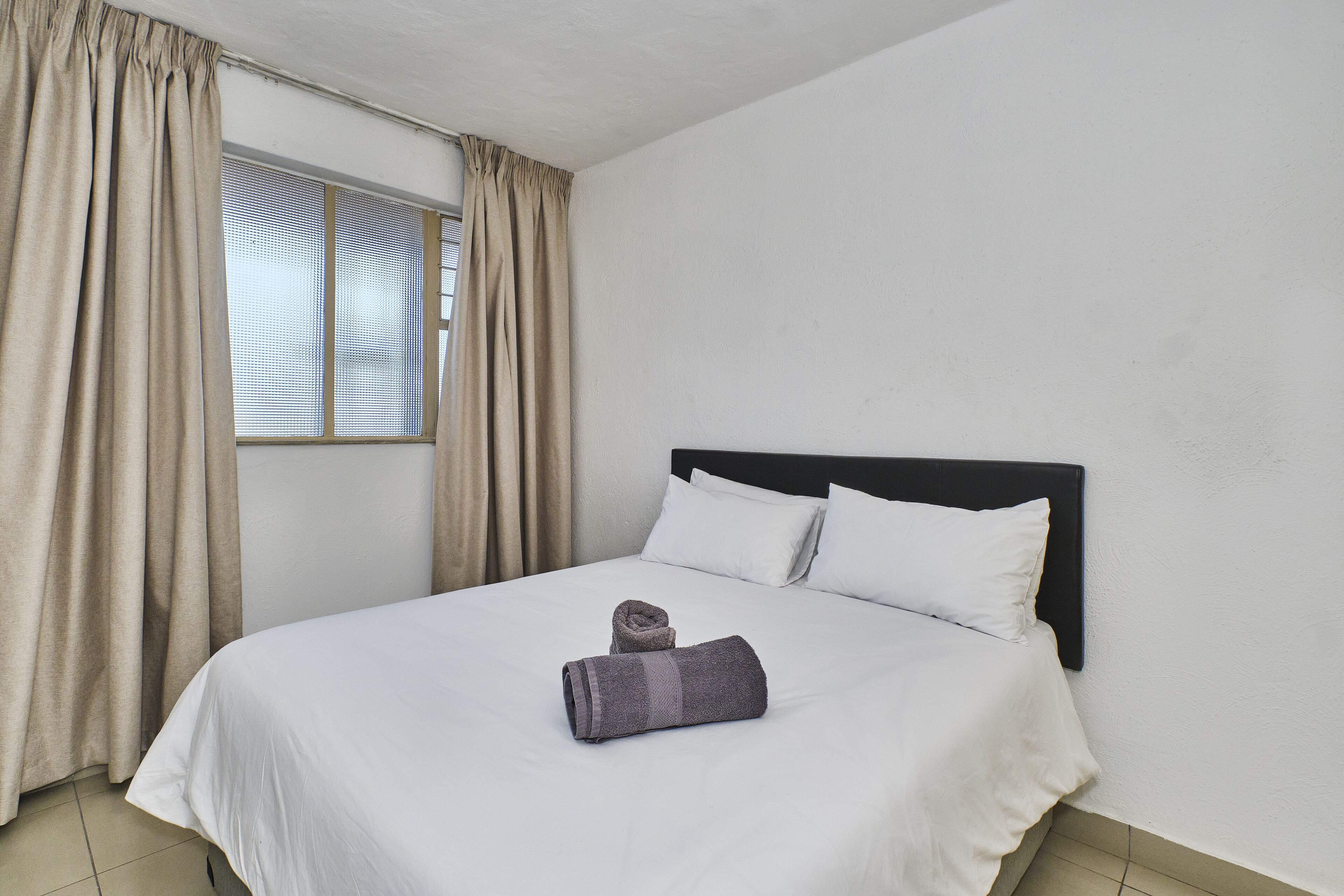 Cama bem arrumada com toalha enrolada, roupa de cama branca e janela com cortinas.