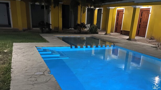 Piscina com borda azul, rodeada por um deck de concreto e um edifício amarelo ao fundo.