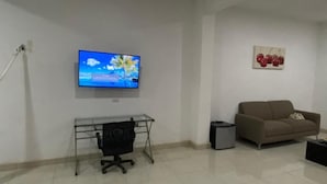 Uma sala de estar com televisão na parede, uma escrivaninha com cadeira, um sofá e uma pequena mesa.