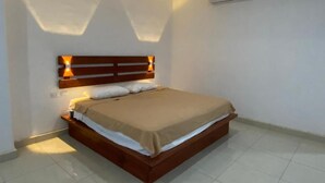 Uma cama de solteiro com cabeceira de madeira e um edredom bege em quarto com paredes brancas e chão de azulejos.