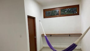 Quarto com uma rede pendurada em uma viga de madeira, uma porta e uma janela com vista para árvores.