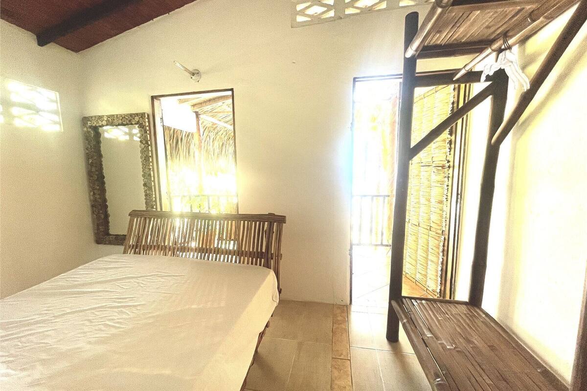 Quarto com cama, um banquinho de madeira, espelho e janela com vista para uma construção de palha.