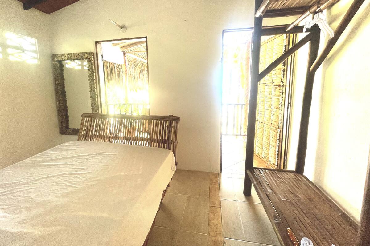 Quarto com cama, um banco de madeira, espelho e janela com vista para uma construção de palha.