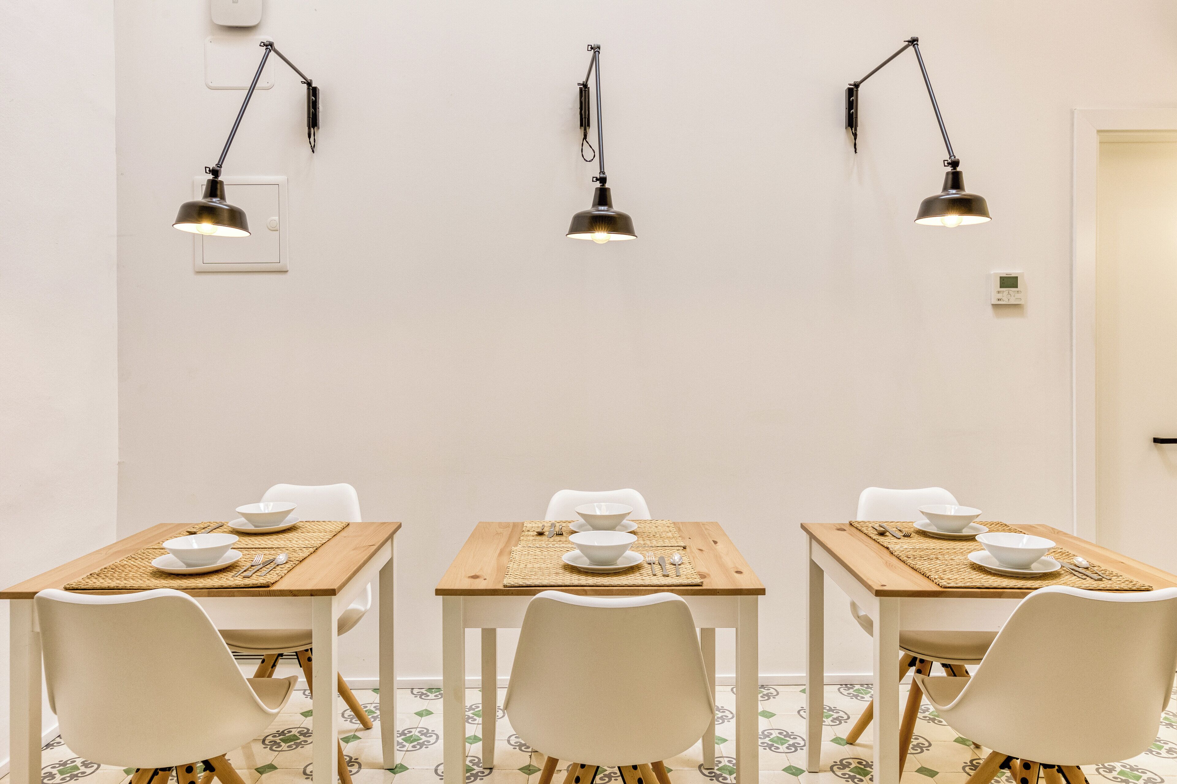 Uma área de refeições com três mesas, cada uma com uma tigela branca, prato e talheres. Três luminárias pendentes pretas estão suspensas acima das mesas.