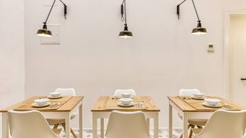 Uma área de refeições com três mesas, cada uma com uma tigela branca, prato e talheres. Três luminárias pendentes pretas estão suspensas acima das mesas.