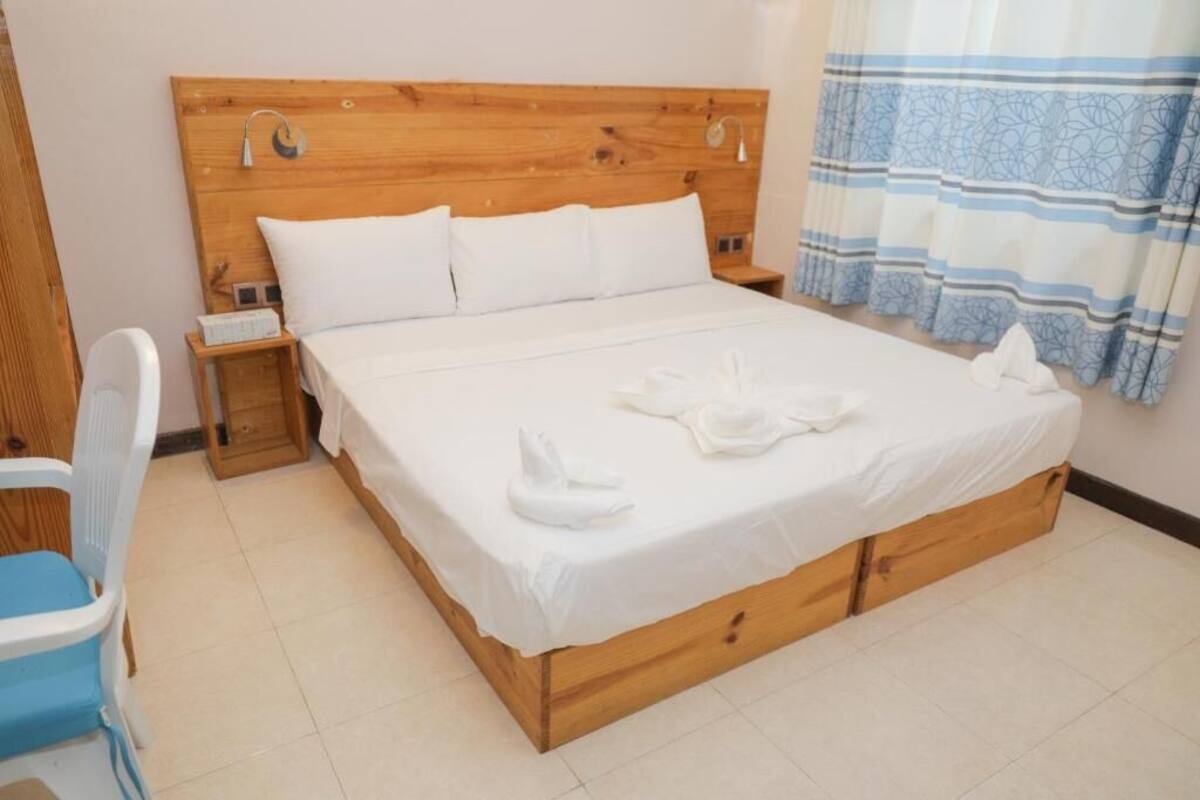 Cama bem arrumada com lençóis e toalhas brancas, cabeceira de madeira, mesa de cabeceira com abajur e cadeira com almofada azul.