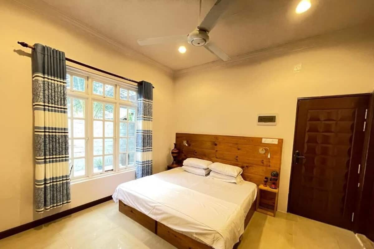 Quarto com cama, cabeceira de madeira, mesa de cabeceira, janela com cortinas e porta.