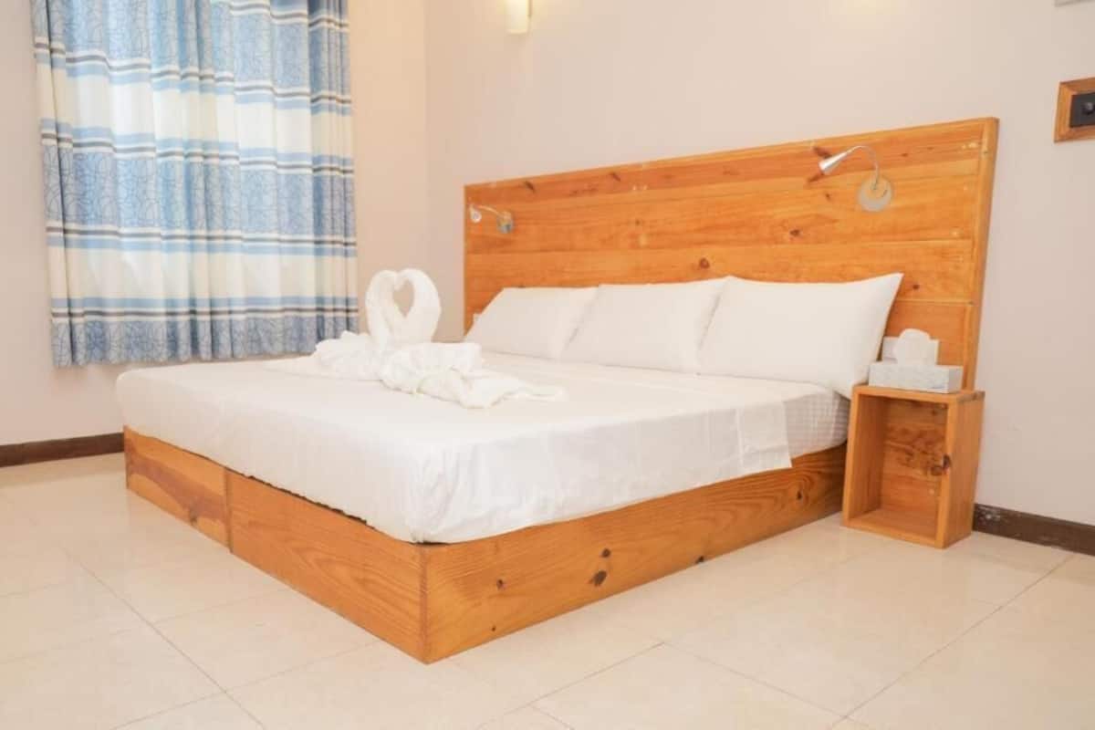 Uma cama bem arrumada com cabeceira de madeira, mesa de cabeceira com caixa de lenços e janela com cortinas listradas.