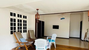 Um quarto amplo com piso de madeira, uma grande janela com vários painéis, um ventilador de teto e uma televisão fixada na parede.