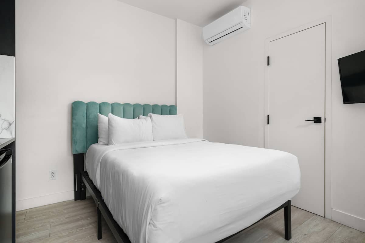 Quarto com cama, parede branca, aparelho de ar condicionado, televisão e porta.
