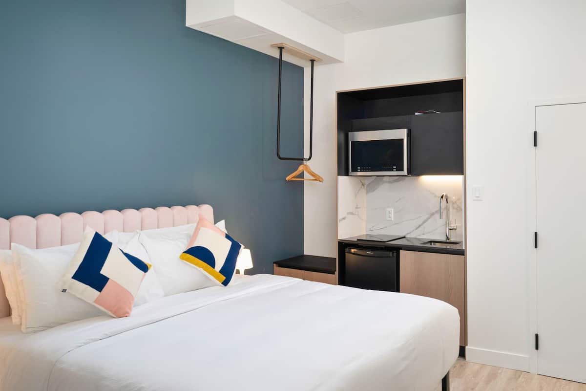 Quarto com cama, mesa de cabeceira e cozinha compacta, incluindo micro-ondas e pia.