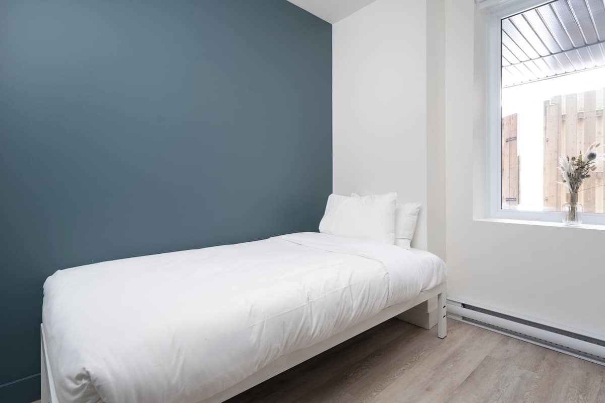 Uma cama de solteiro em um quarto com parede de destaque verde-água e janela com vista para uma cerca de madeira.
