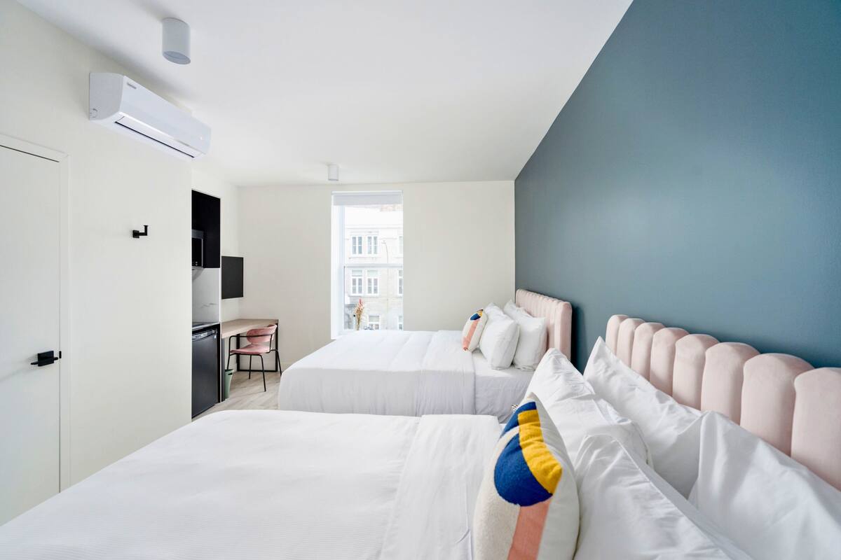 Quarto de hotel moderno com duas camas, uma escrivaninha e vista para a cidade.